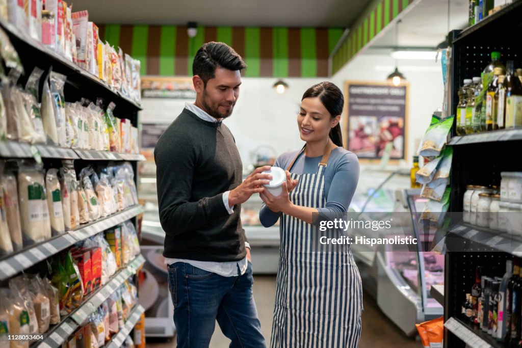 Latin American Sales Clerk At A Supermarket Suggesting A Product To Male Customer Smiling High