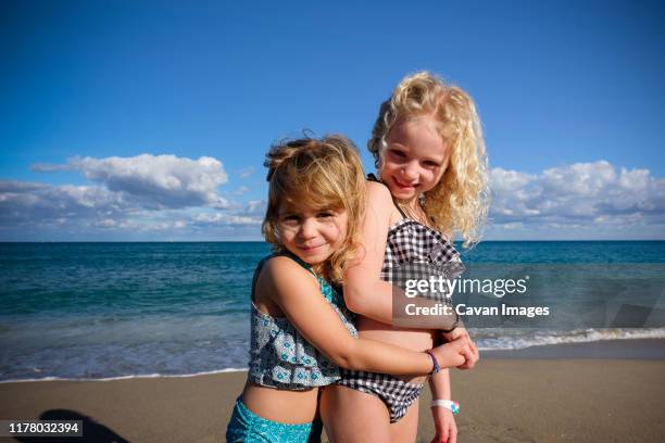 Two Girls Hugging Standing On The Beach In Bathing Suits On Sunny Day