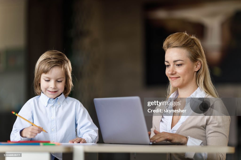Working Single Mother Using Laptop While Her Son Is Coloring At Home