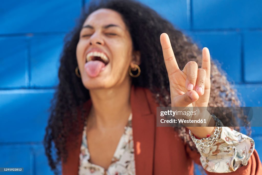 Young Woman Sticking Out Tongue Doing Horn Sign Against Blue Wall High