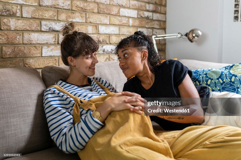 Smiling Lesbian Couple Sitting With Hands Clasped On Sofa At Home High
