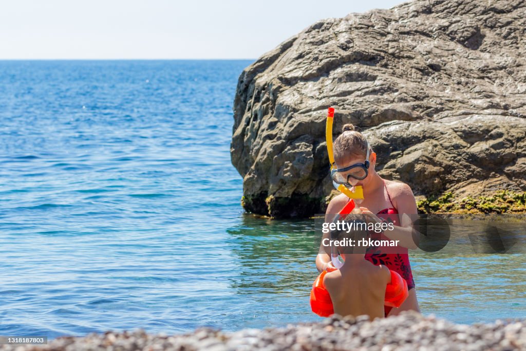 Adult Sister Helping Brother Putting On Scuba Mask And Snorkel During Summer Vacation At Sea