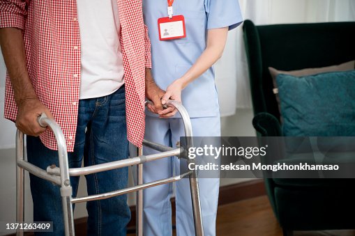 Female Caregiver Assisting Elderly Man To Walk With Frame At Home