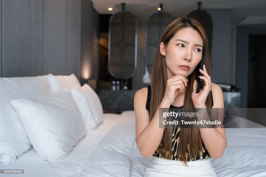Asian Woman Making A Call Using A Phone Sitting On The Bed In The Hotel