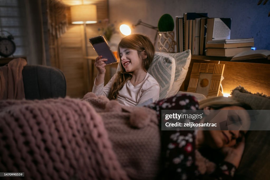 Mother And Daughter Sleeping In Bed Together And Girl Using A Phone