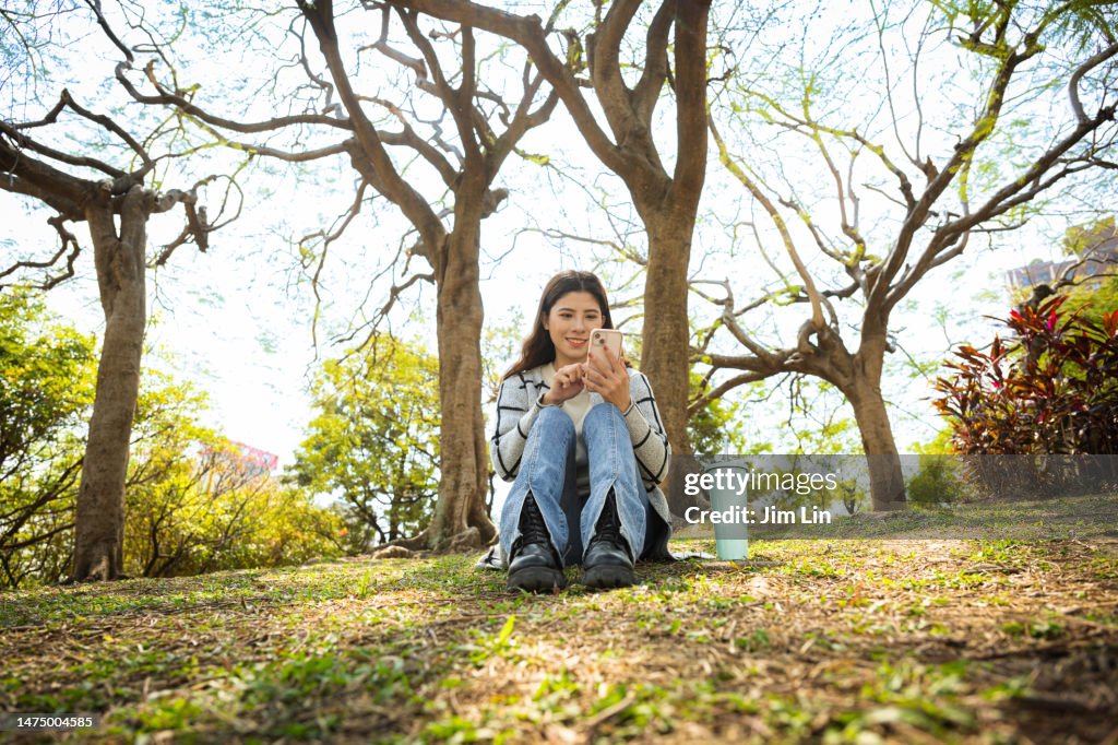 A Young Lady Sitting On Grass At A City Park Using Her Smart Phone As Working Device High Res