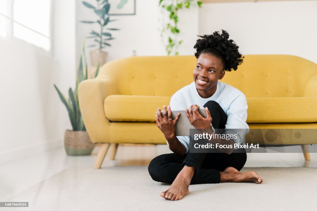 Young Woman Sitting On The Floor In Her Living Room Holding A Digital