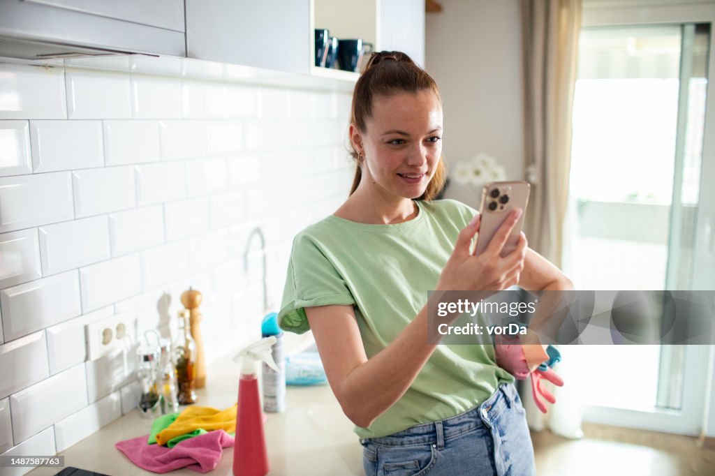 Young Woman Using A Smart Phone While Cleaning The Kitchen In Her Home