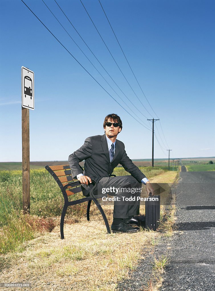 Businessman Sitting On Bench At Bus Stop With Suitcase High Res Stock