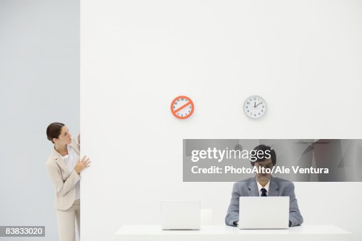 Female Office Worker Peeking Around Corner At Clocks On Wall Colleague