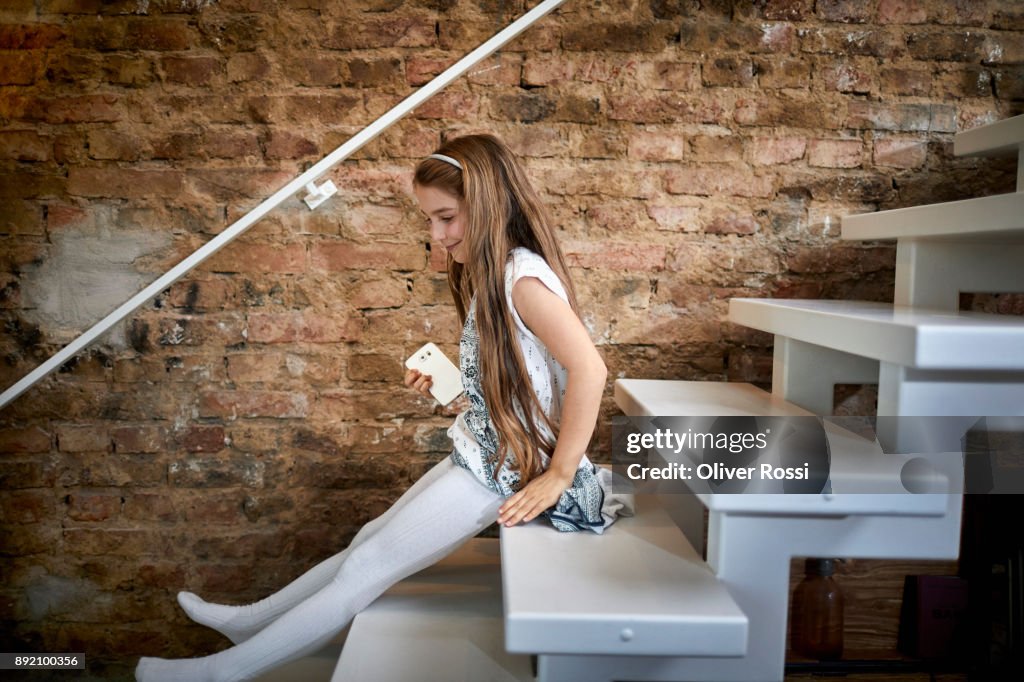 Smiling Girl Holding Cell Phone Sitting On Stairs At Home High Res
