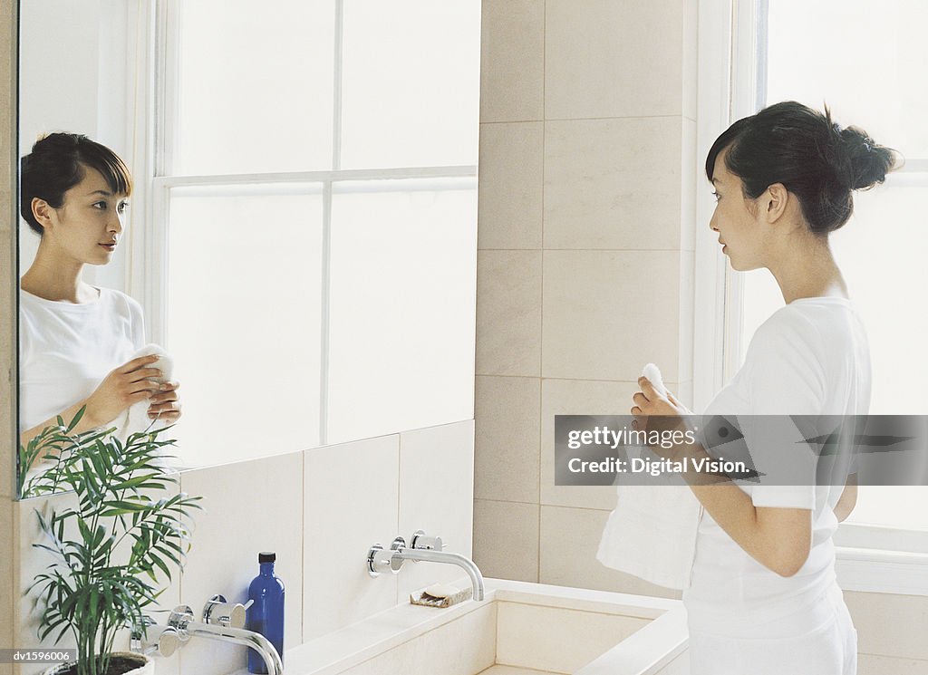 Woman Stands Looking At Herself In A Bathroom Mirror Holding A Towel